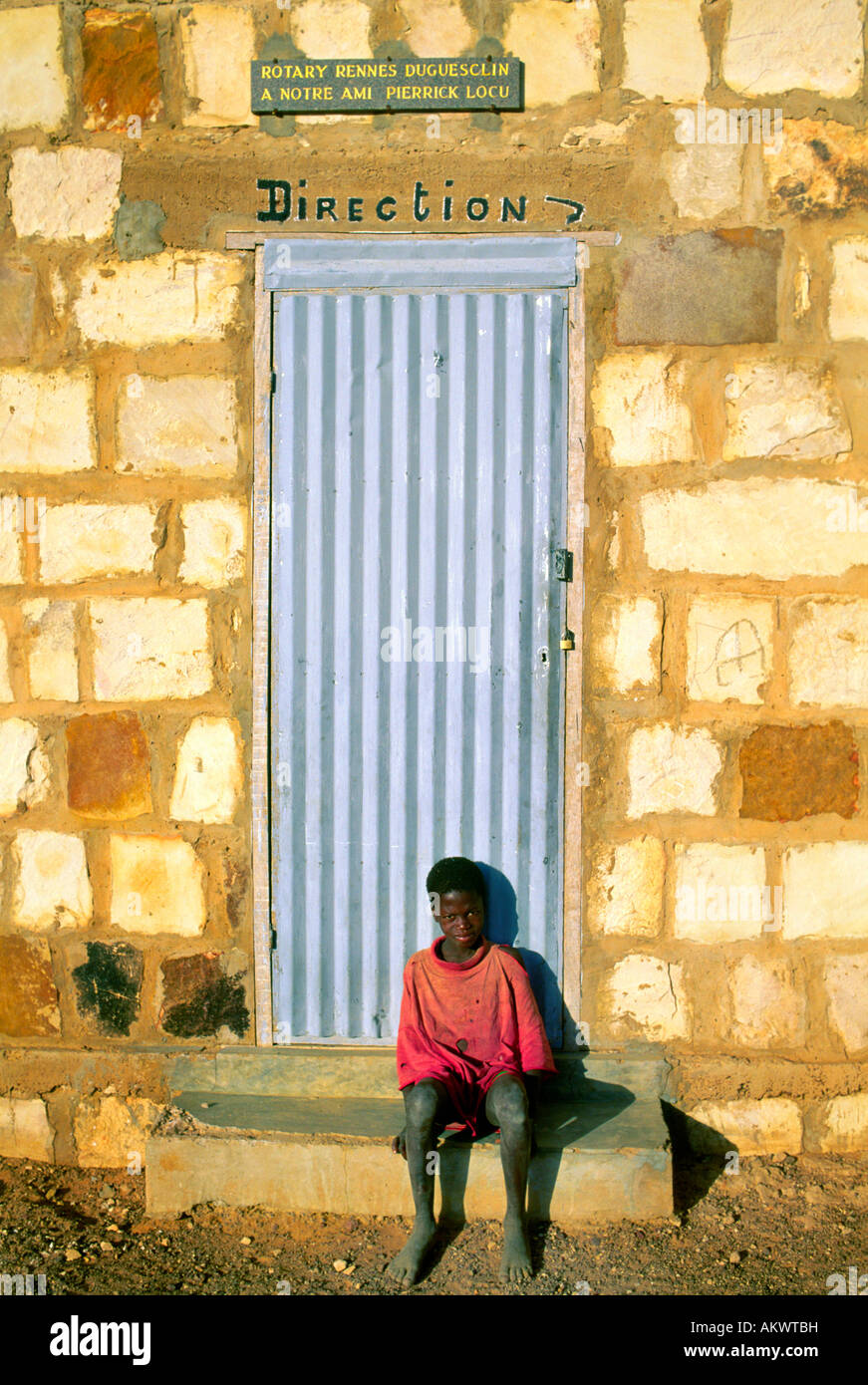 A Dogon village boy sits on the step in front of a stone building built ...