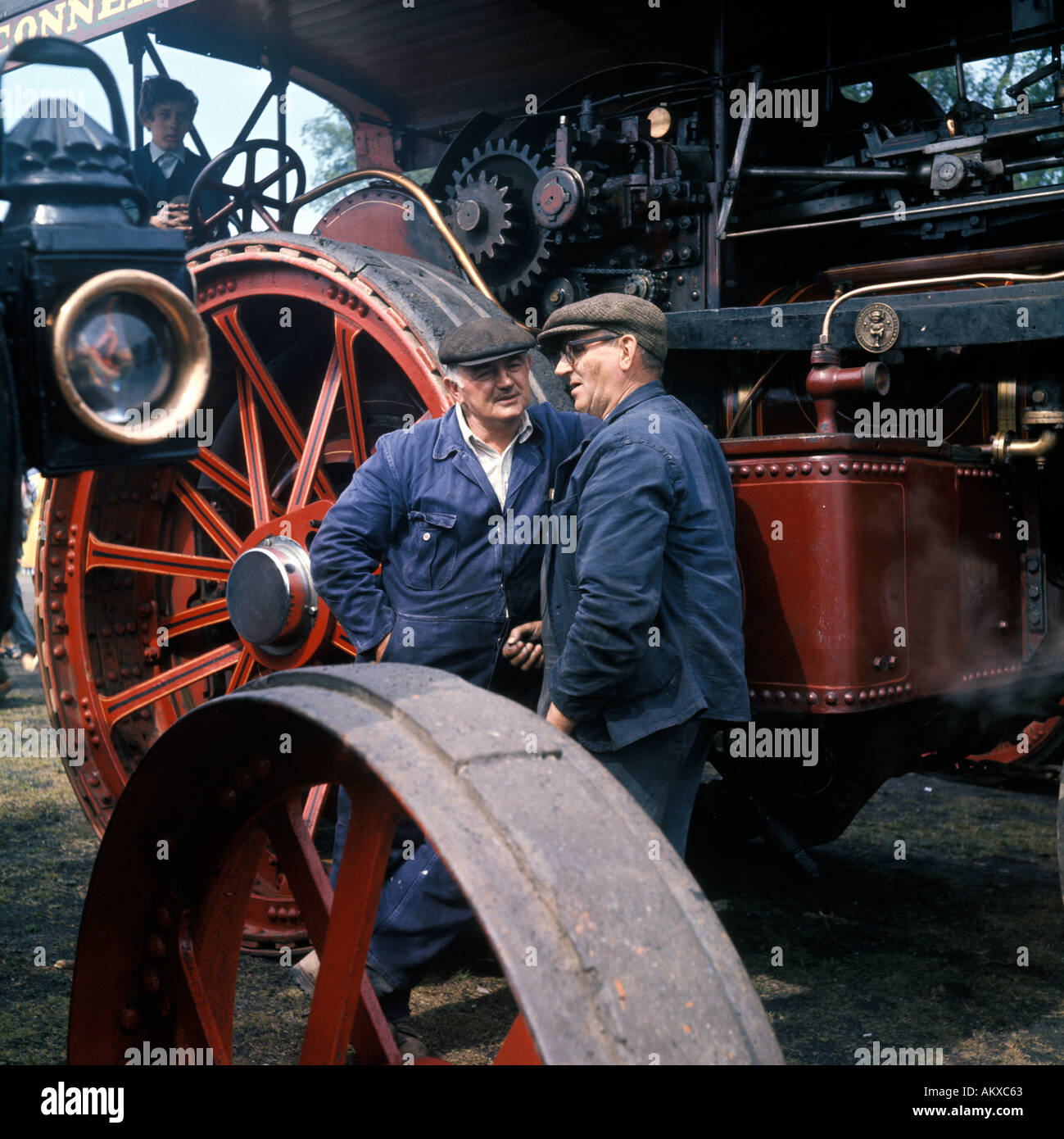 Two men in boilersuits and cloth caps in conversation leaning against ...