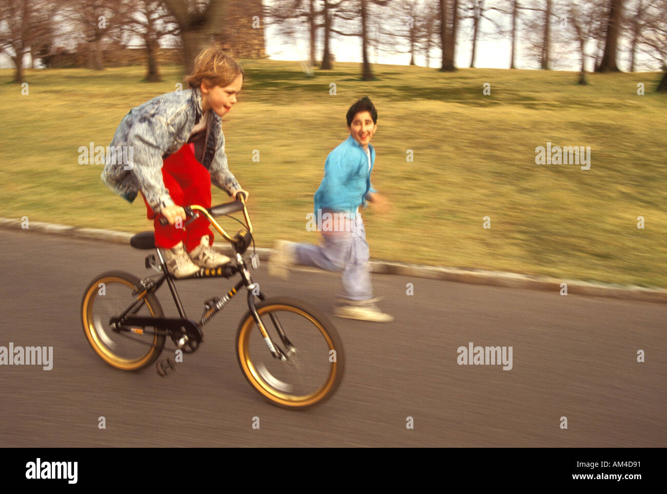 boys ride bikes. One does a stunt by standing on the frame of his ...
