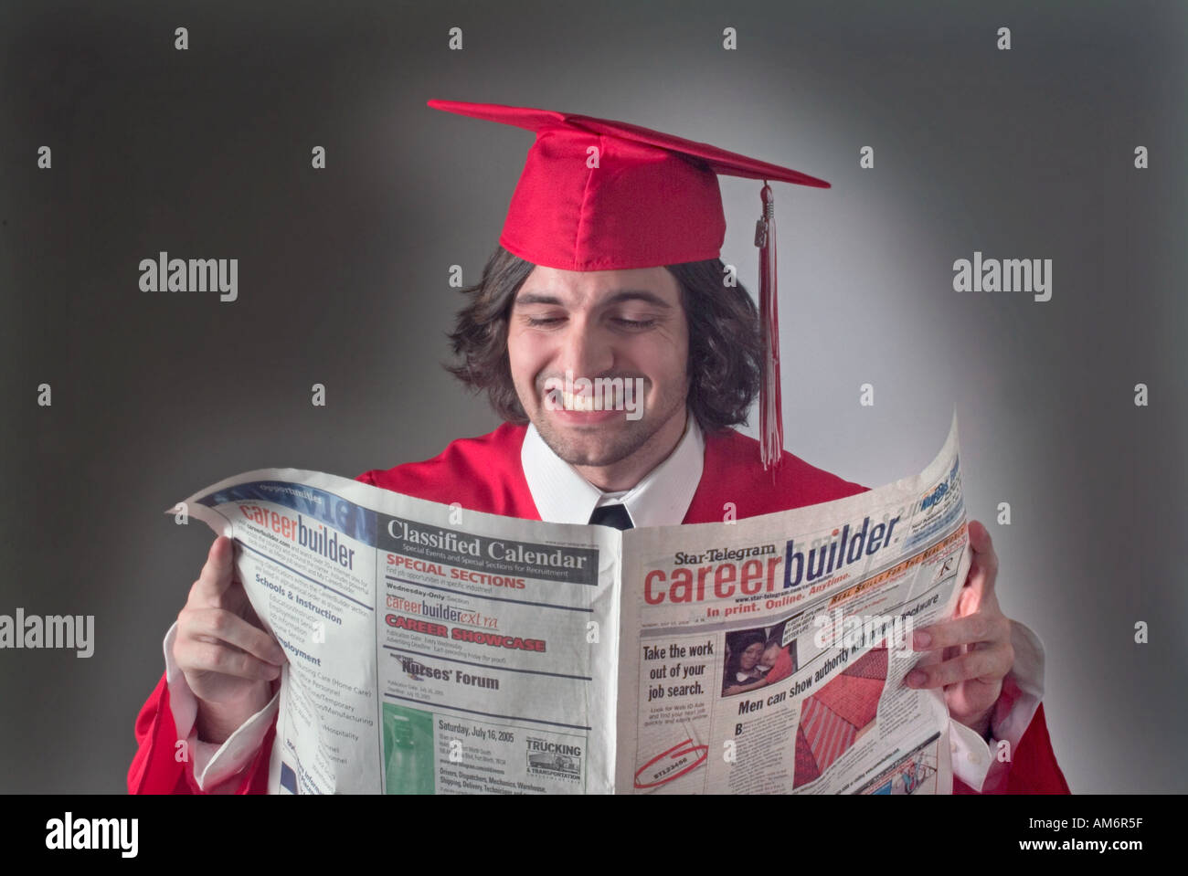 Worried male graduate in cap and gown reading want ads in newspaper ...