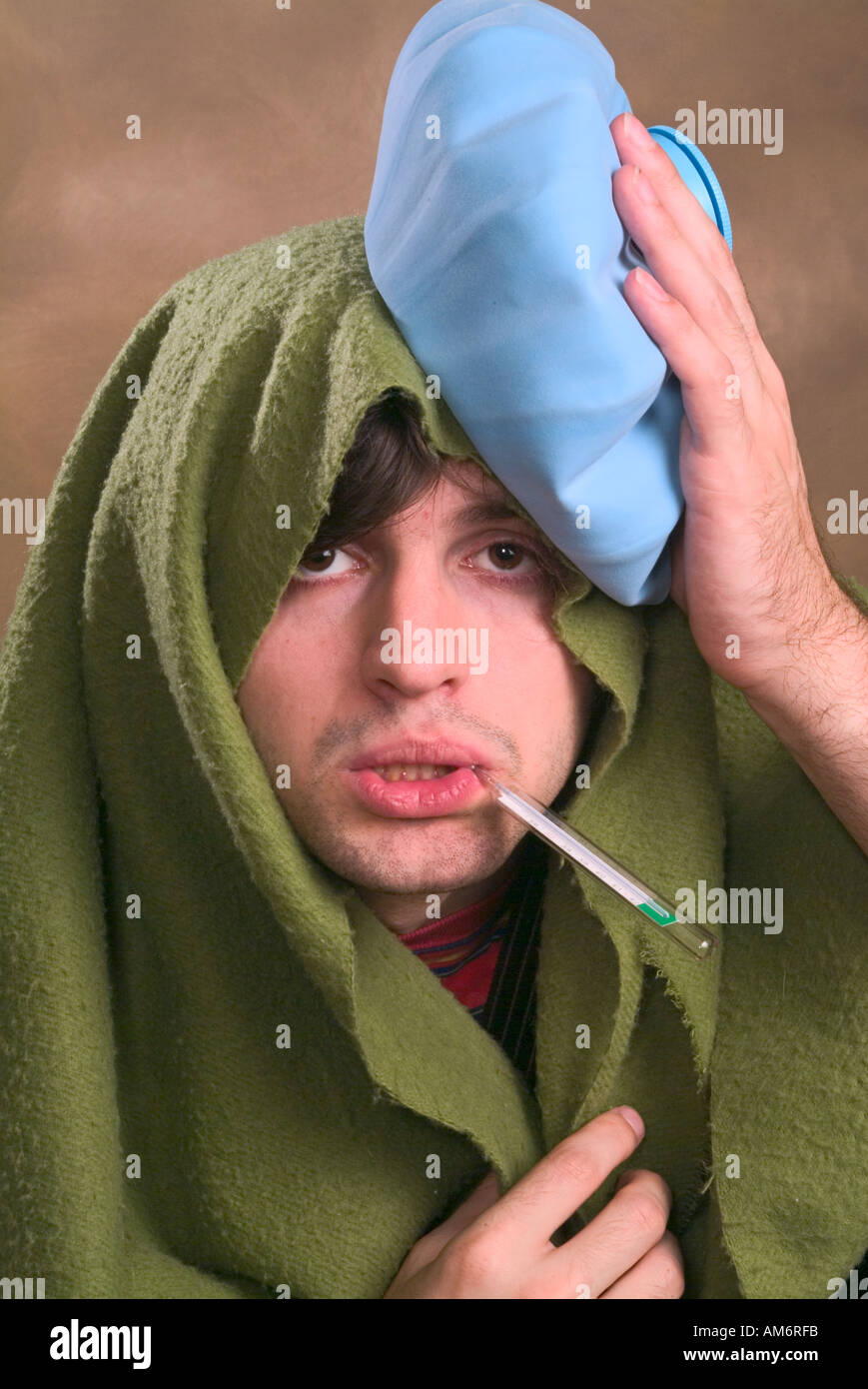 Sick male wrapped in blanket with thermometer and ice pack on head ...