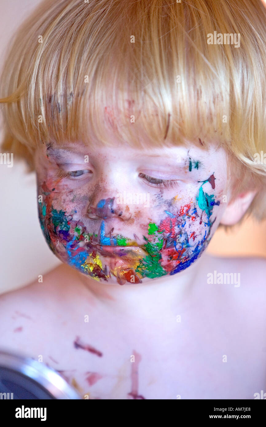Cheeky young child with face covered in colourful paint looking in a ...