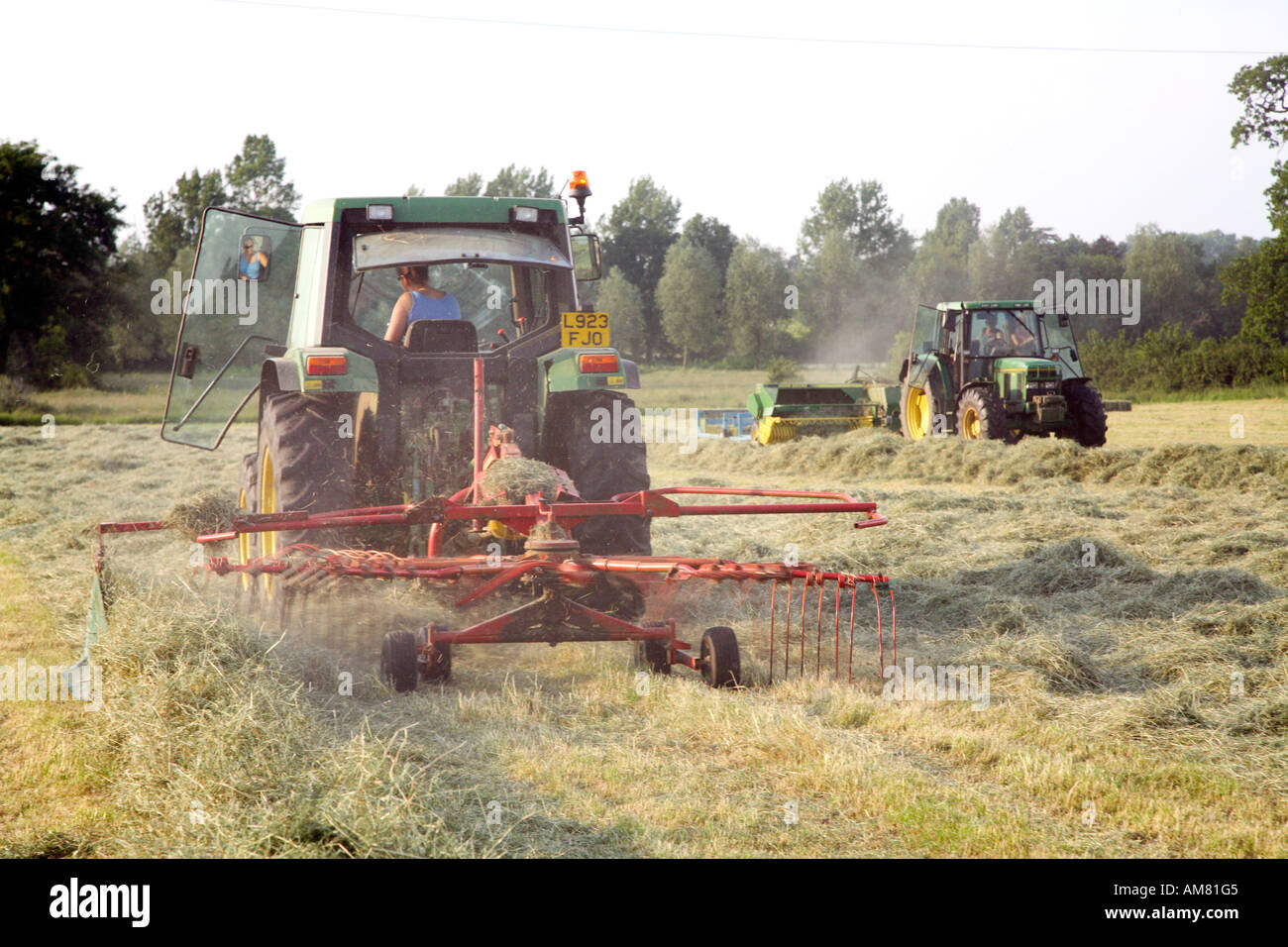 Female farm worker raking hay into rows with tractor with baler at work ...