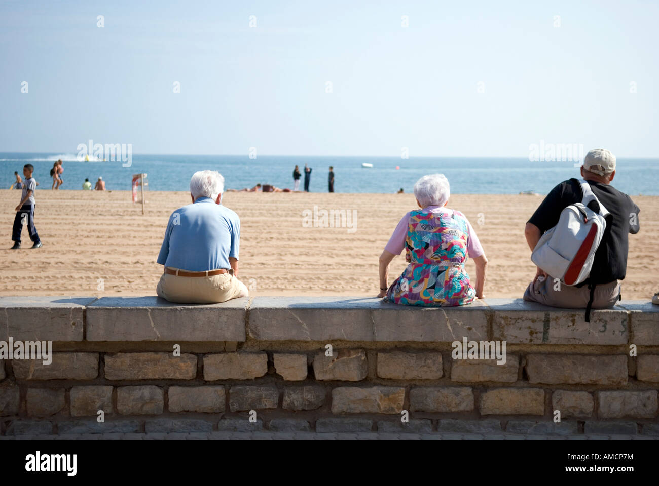 a rear view of three old people sitting on wall overlooking a beach in ...