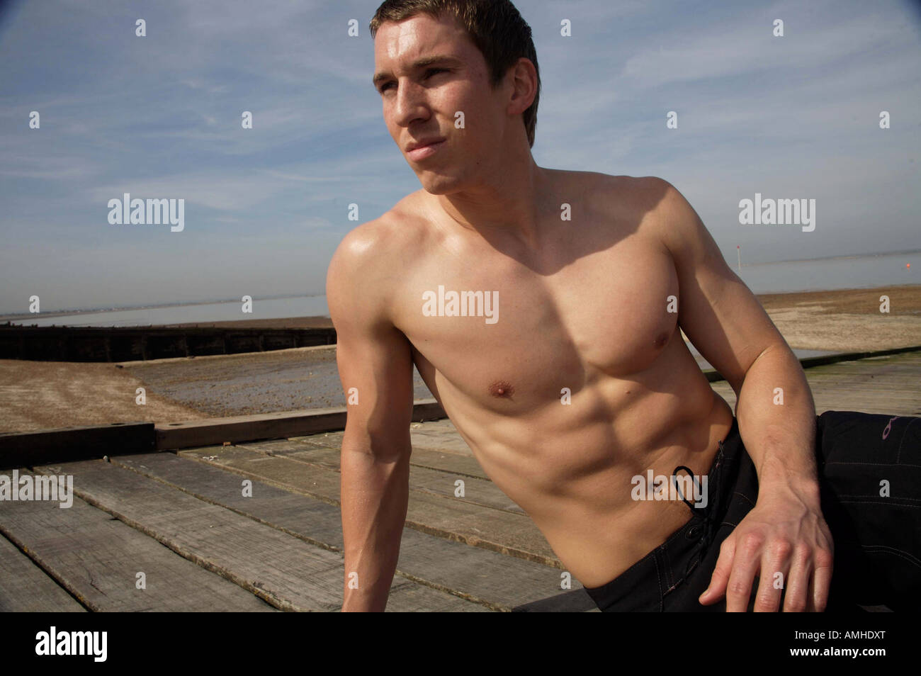 Young, healthy, well-built man sits on the jetty showing his muscles ...