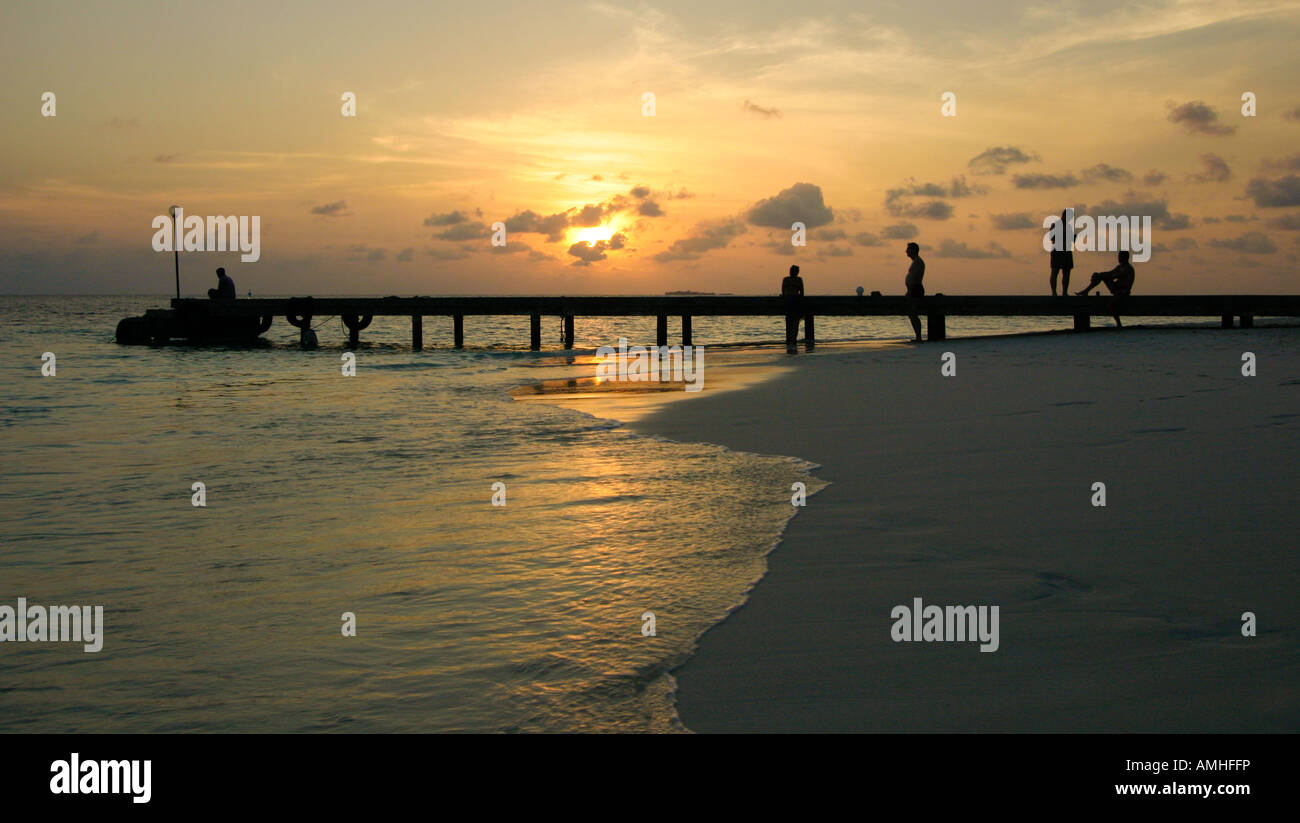 Silhouetted figures watch the sun set from the pier of an idyllic beach ...