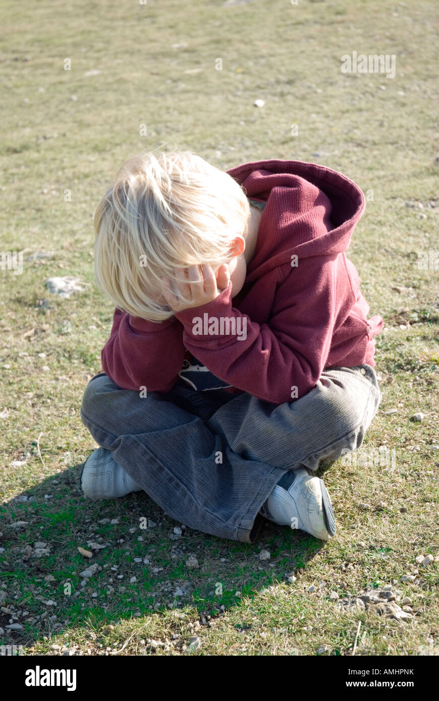 A 6 year old blonde boy sits down on the grass sulking with his head in ...