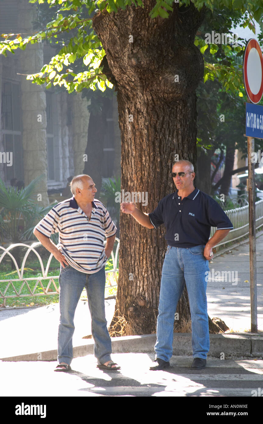 Two men standing talking discussing on a street corner under a big tree ...