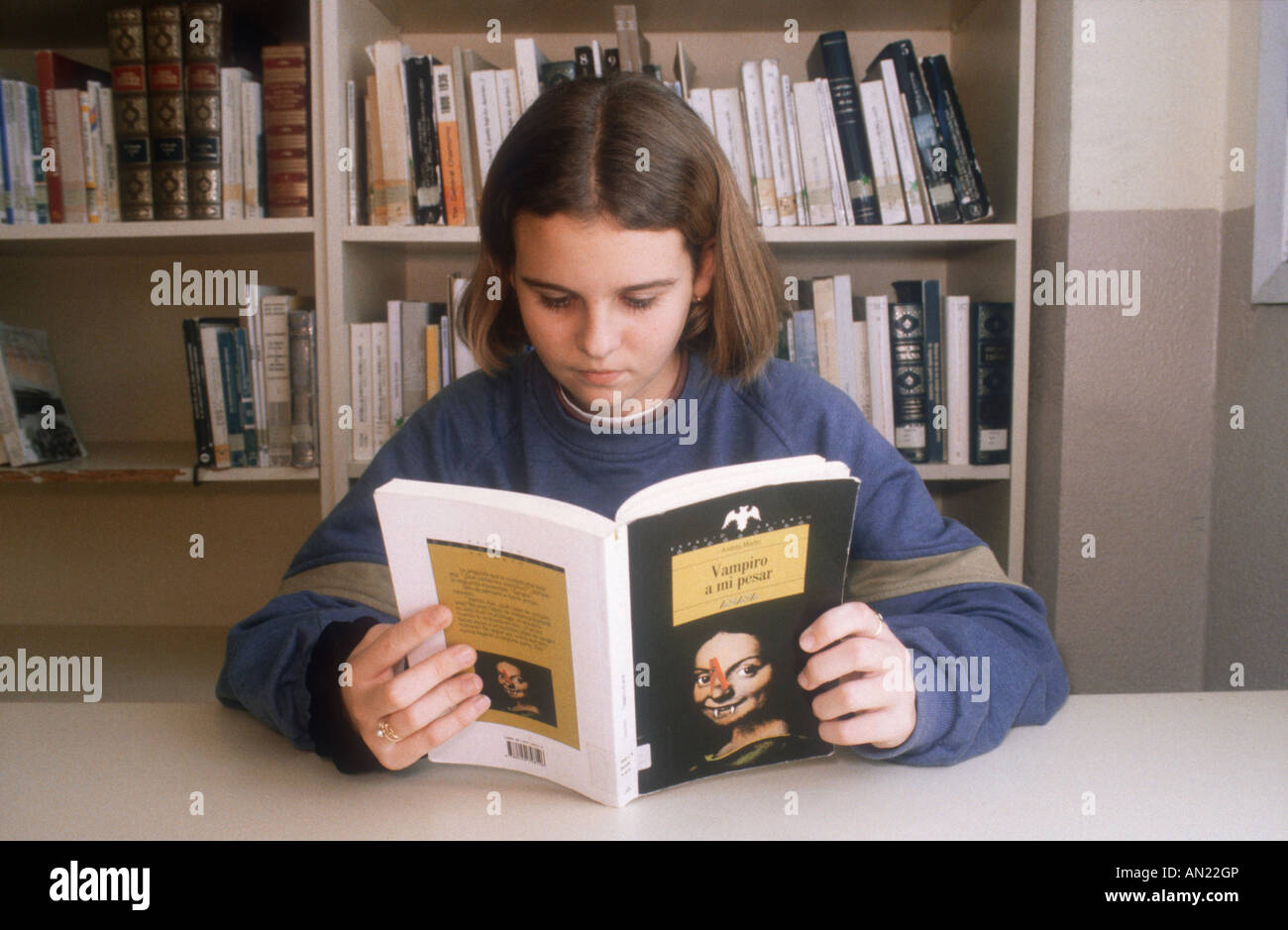 Young girl reading book in school library with bookcase behind her ...