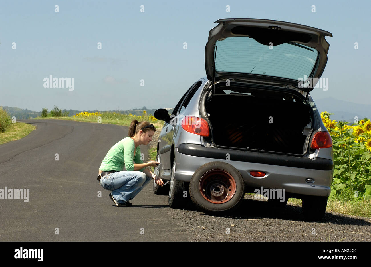 A woman is kneeling beside a car on a rural road, working on a flat ...