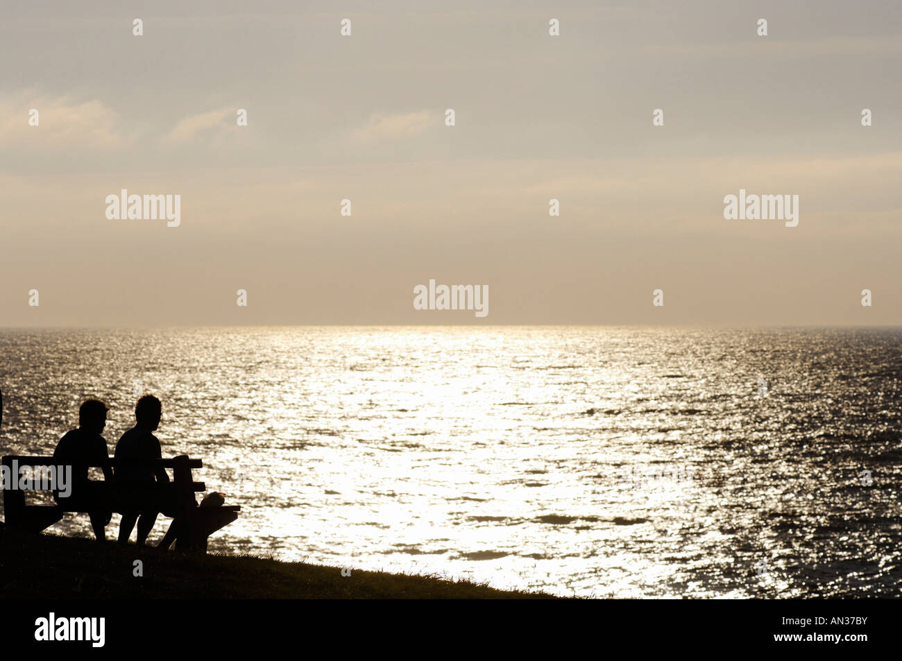 Two people sitting on bench watching the sun set over Cornwall Stock ...