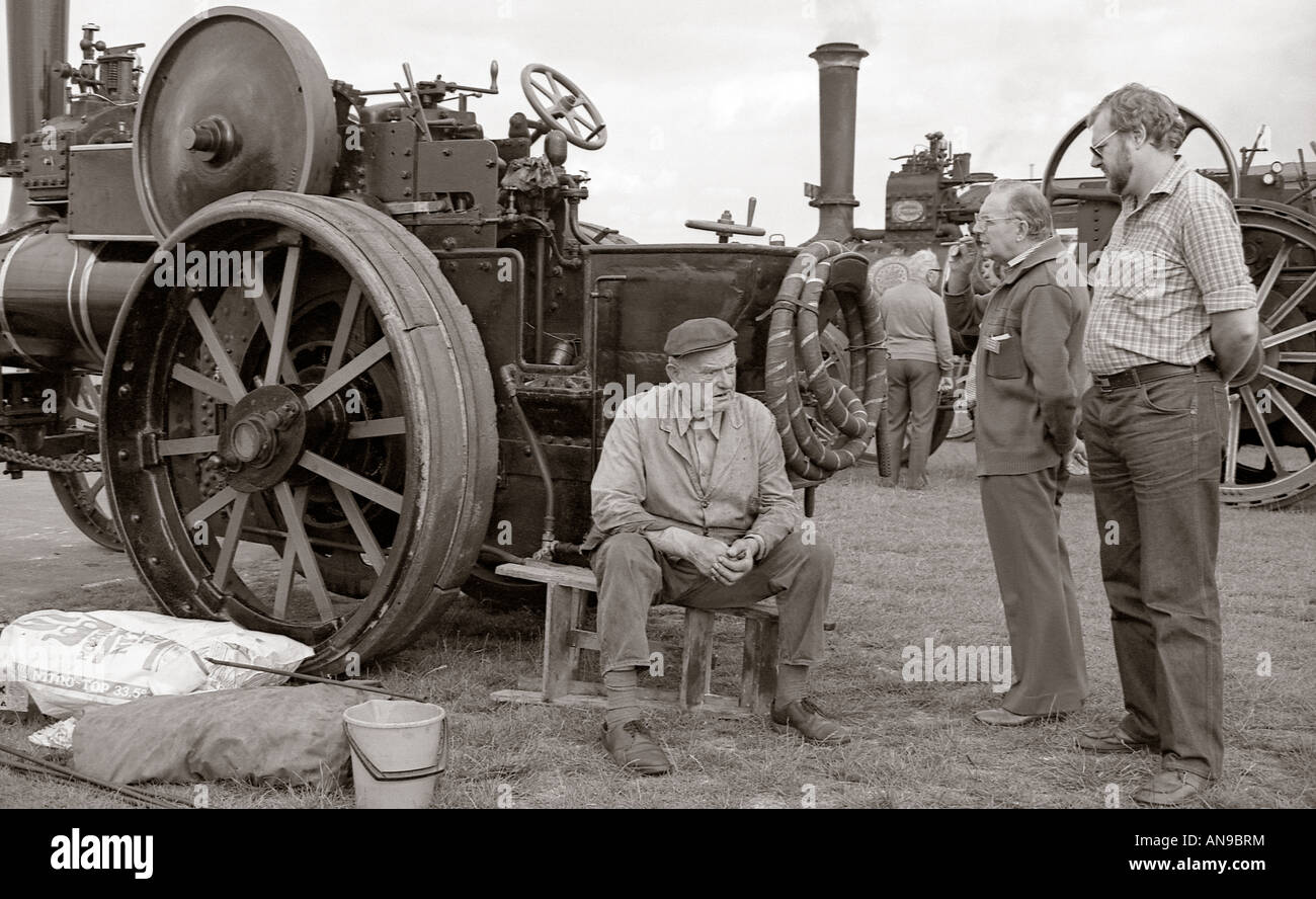Three men at traction engine rally with engine owner sitting down in ...