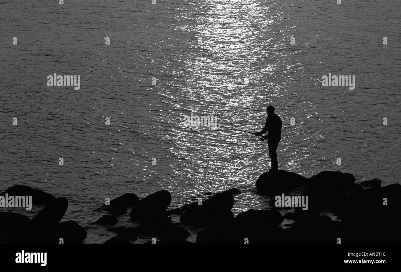 Sea angler fishing from rocks with bright sun reflecting on the water ...