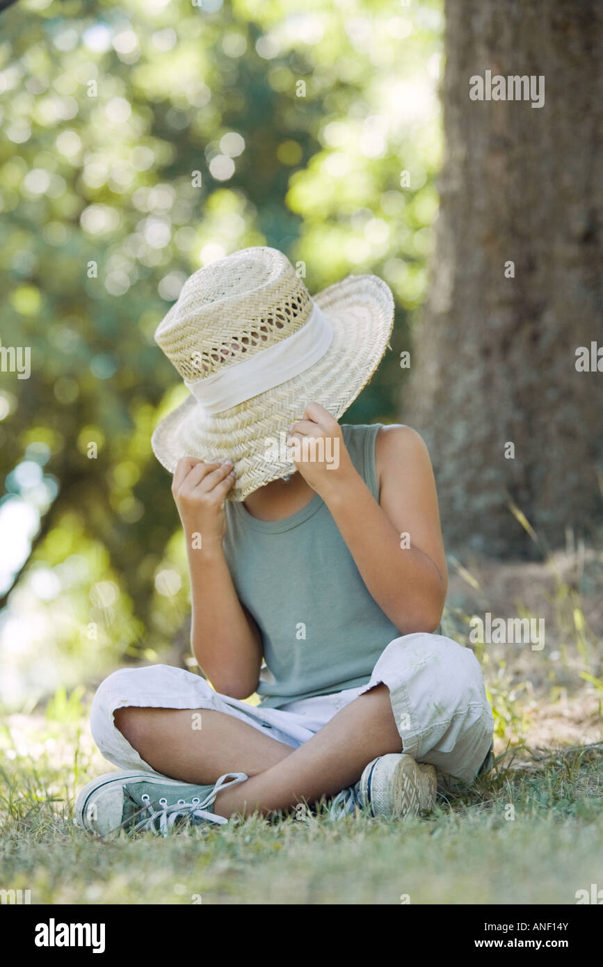 Boy sitting outdoors, pulling straw hat down over face Stock Photo - Alamy