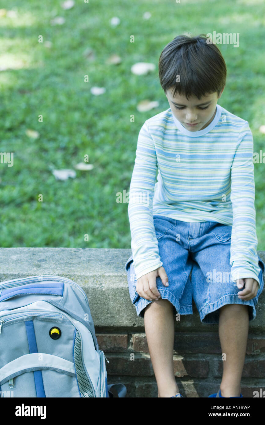 Boy sitting on low wall, backpack by side, looking down Stock Photo - Alamy