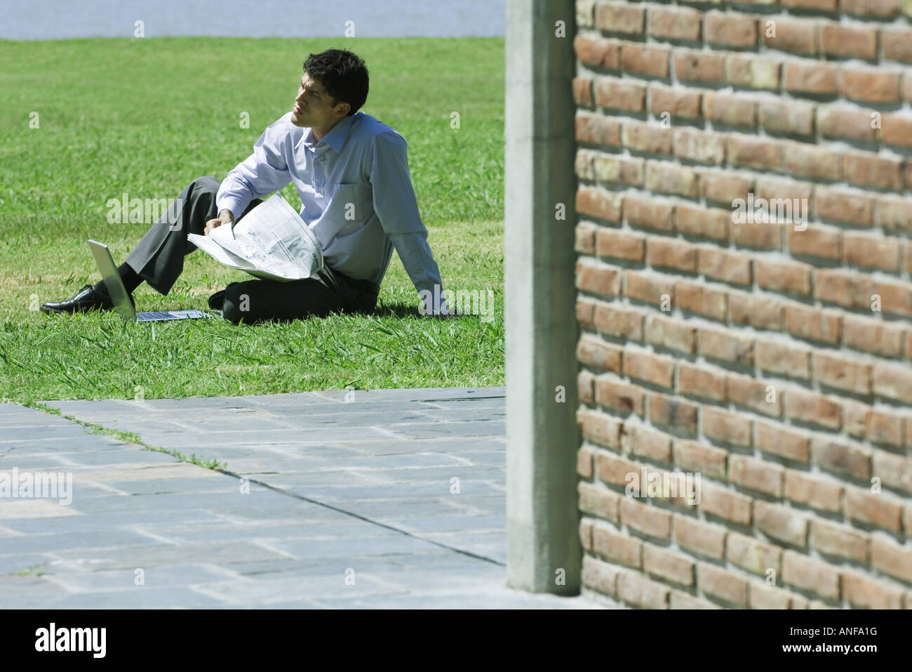 Businessman sitting on grass with laptop and newspaper, corner of brick ...