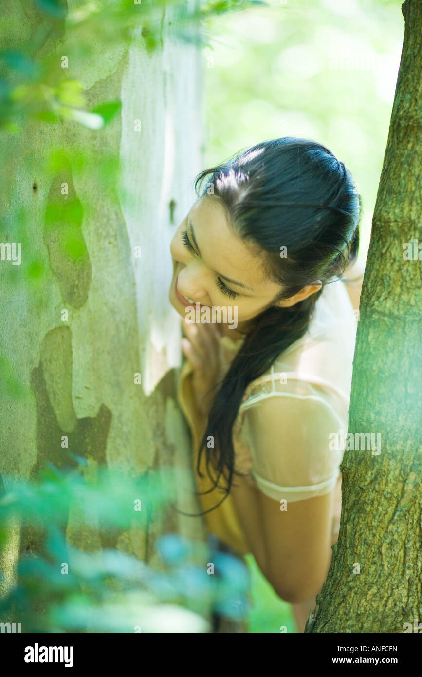 Young woman peeking around tree trunk, side view Stock Photo - Alamy