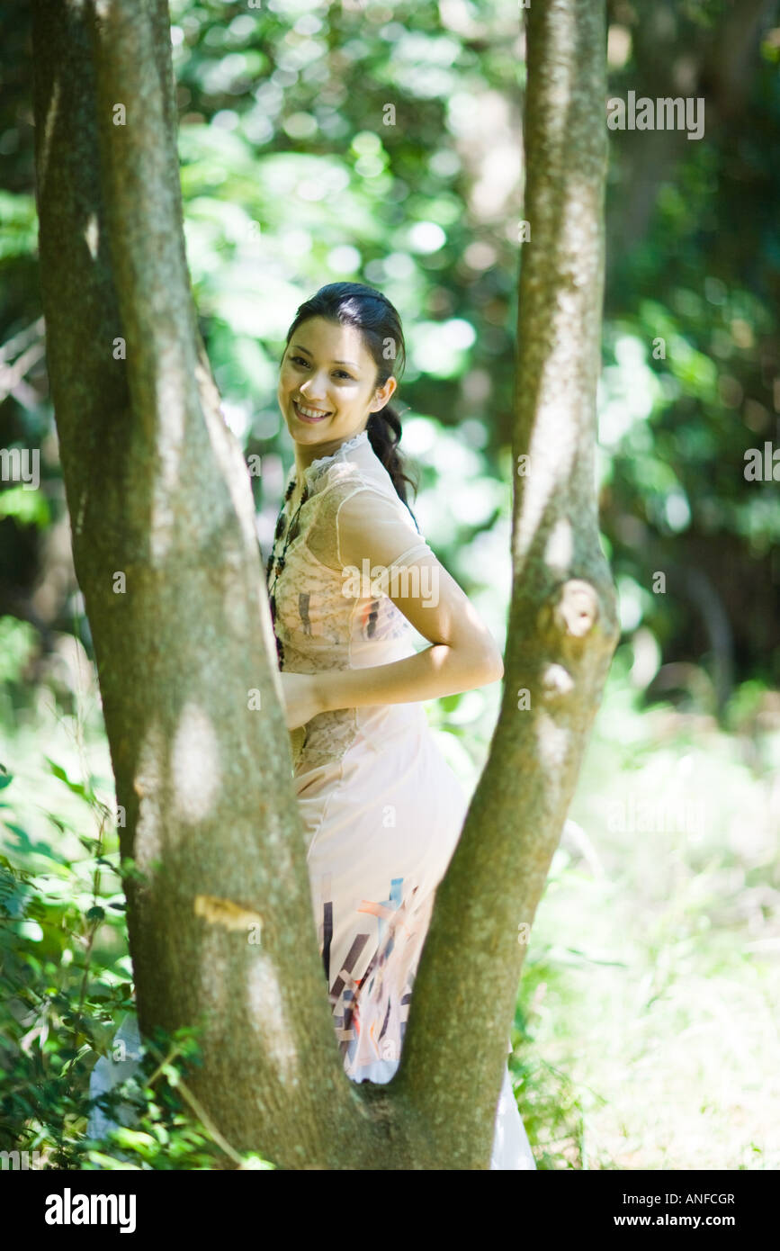 Young woman standing behind tree, smiling at camera Stock Photo - Alamy