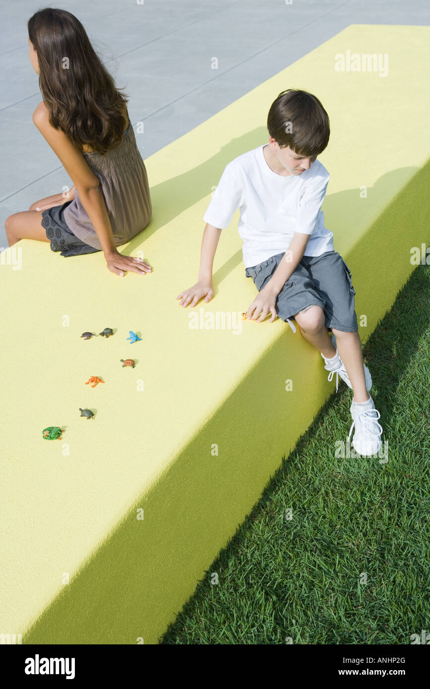 Brother and sister sitting back to back on low wall next to toys, high ...