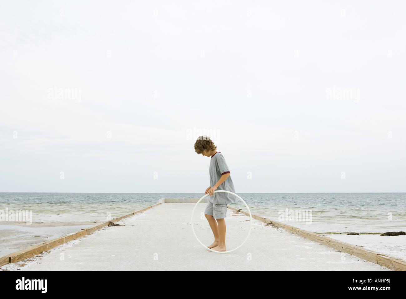 Boy standing on pier holding plastic hoop, looking down, side view ...