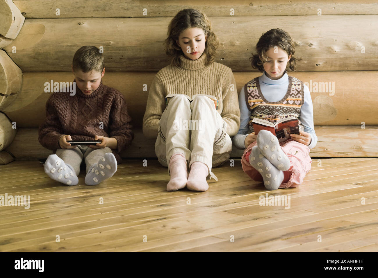 Three young siblings sitting side by side, reading and playing video ...