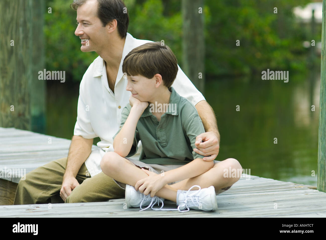 Man sitting on dock with arm around son's shoulder, both looking away ...