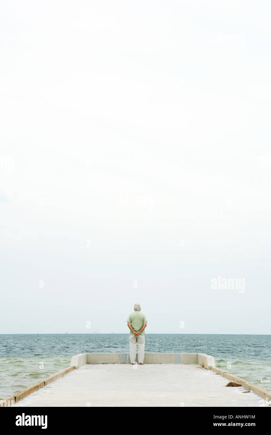 Senior man standing at end of pier, facing ocean, in distance, rear ...