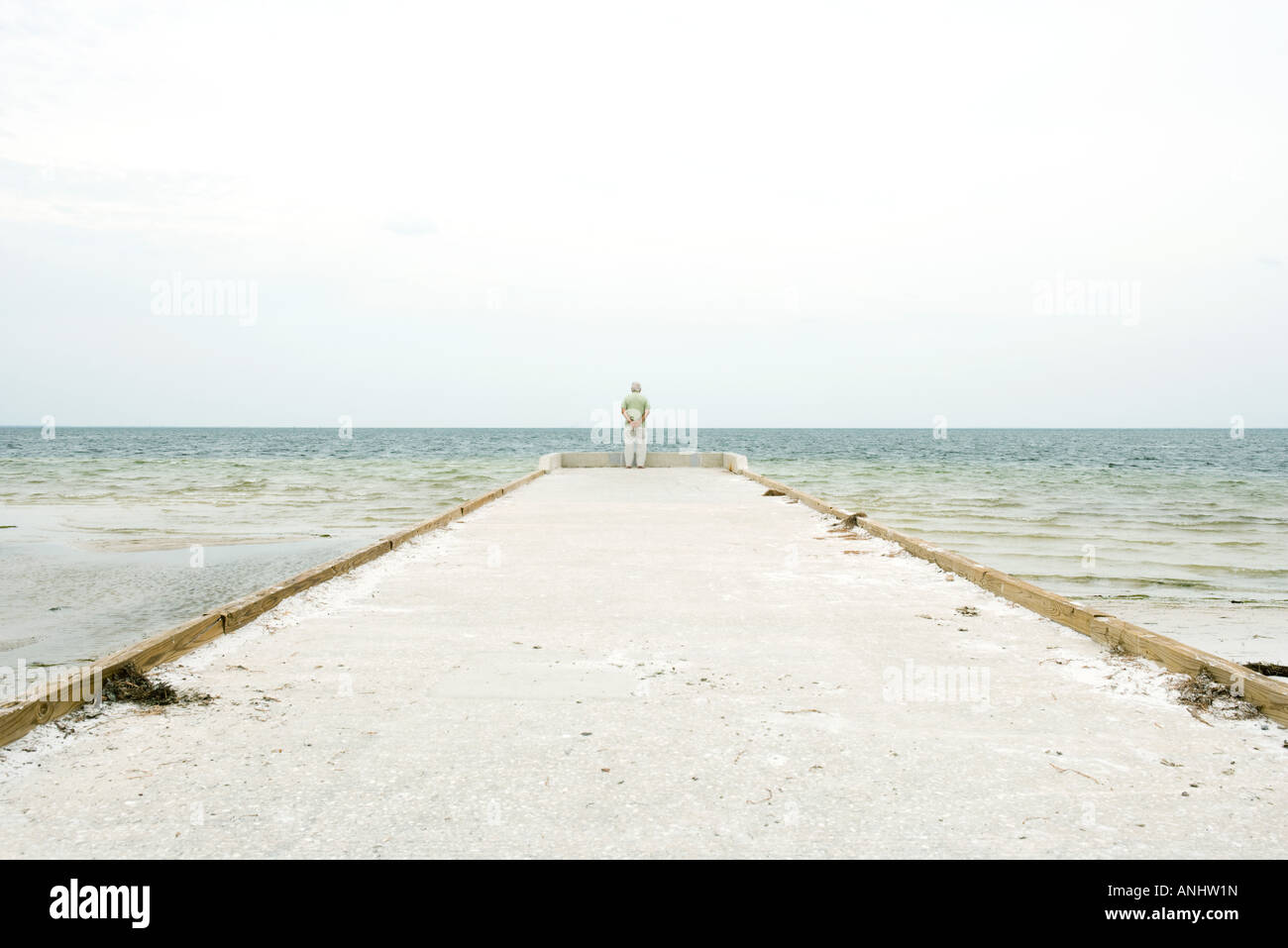 Senior man standing at end of pier, facing ocean, in distance, rear ...