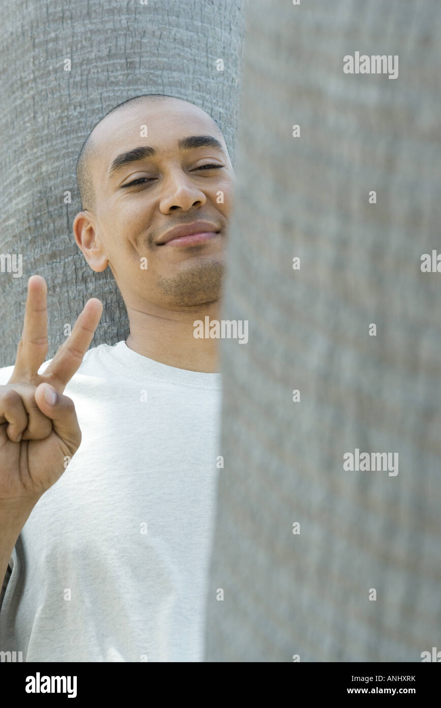 Man leaning against tree trunk, making peace sign, smiling at camera ...