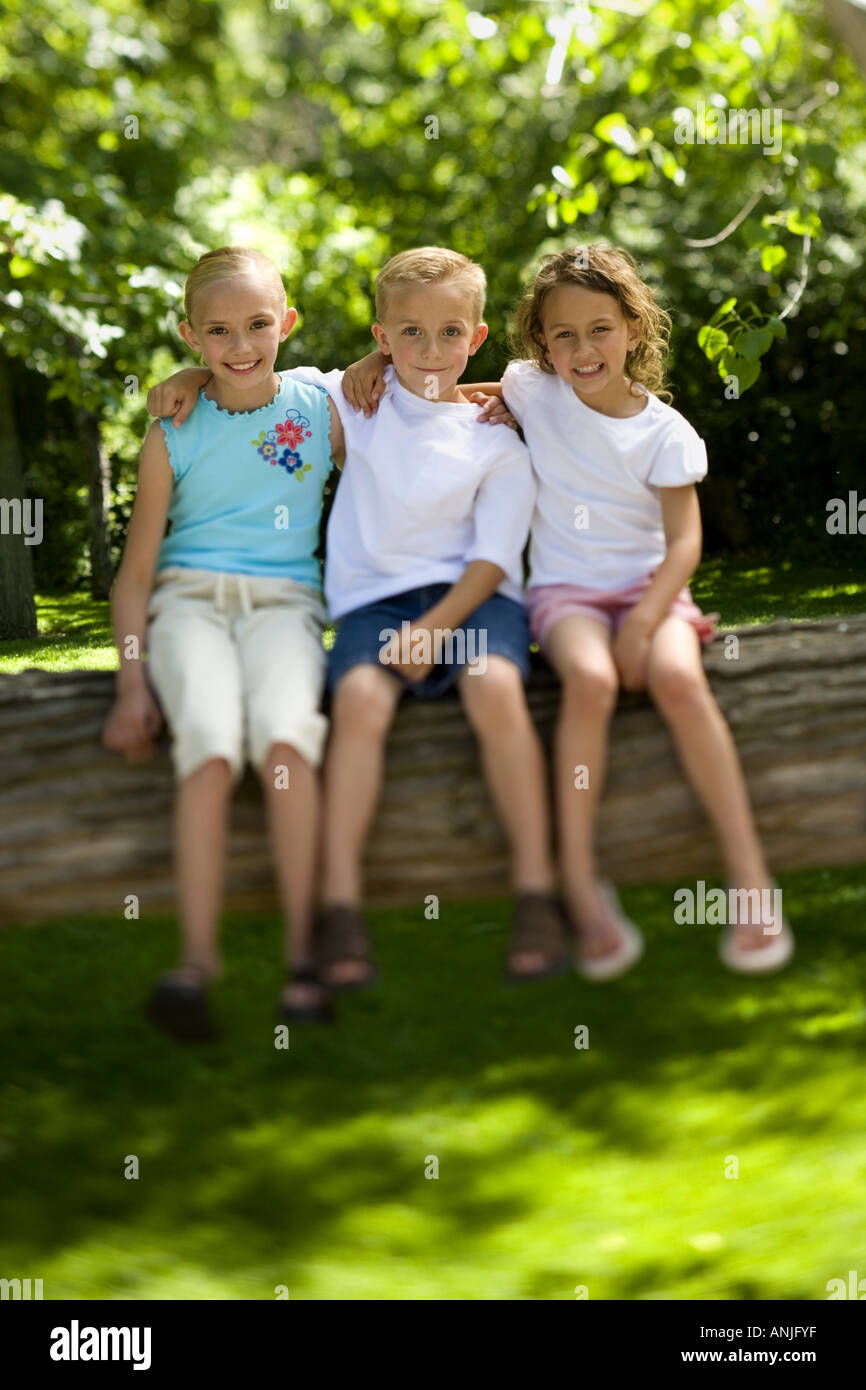 Portrait of two girls and a boy sitting on a tree trunk Stock Photo - Alamy