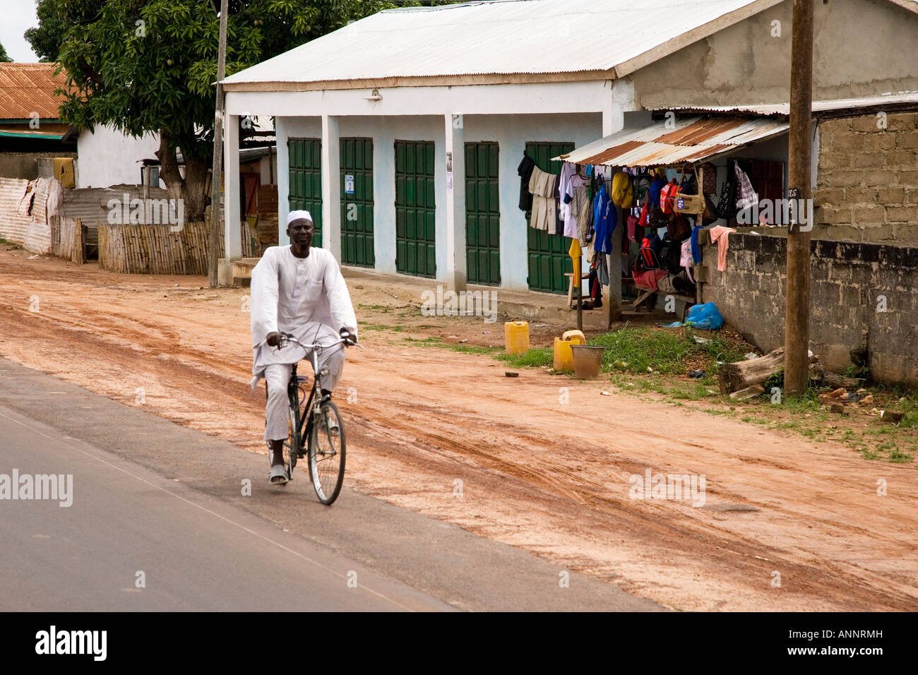 Gambian man in traditional muslim dress riding a bicycle along the road ...