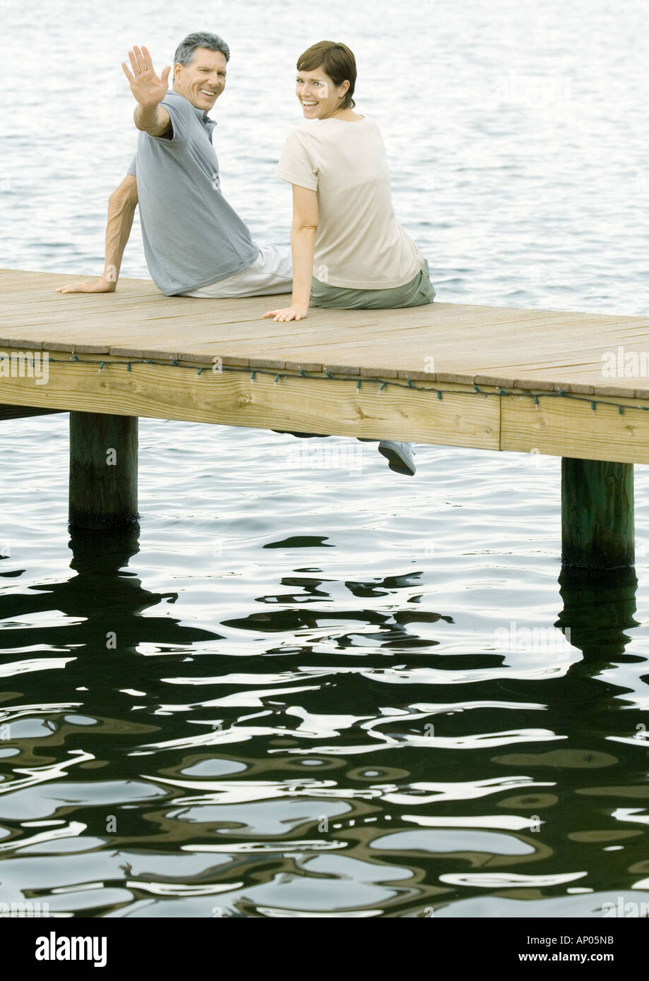 Couple sitting on dock, man waving at camera Stock Photo - Alamy