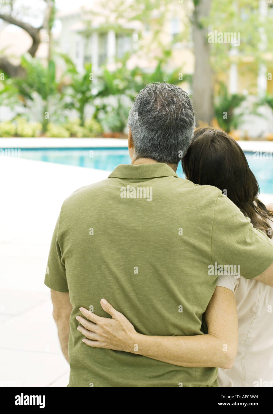 Couple standing with arms around each other, looking at swimming pool ...