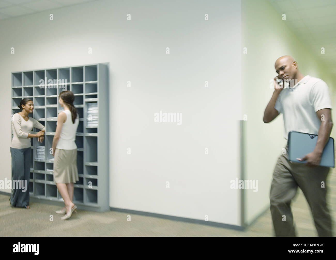 Office scene, two women talking by mailboxes while man using cell phone ...