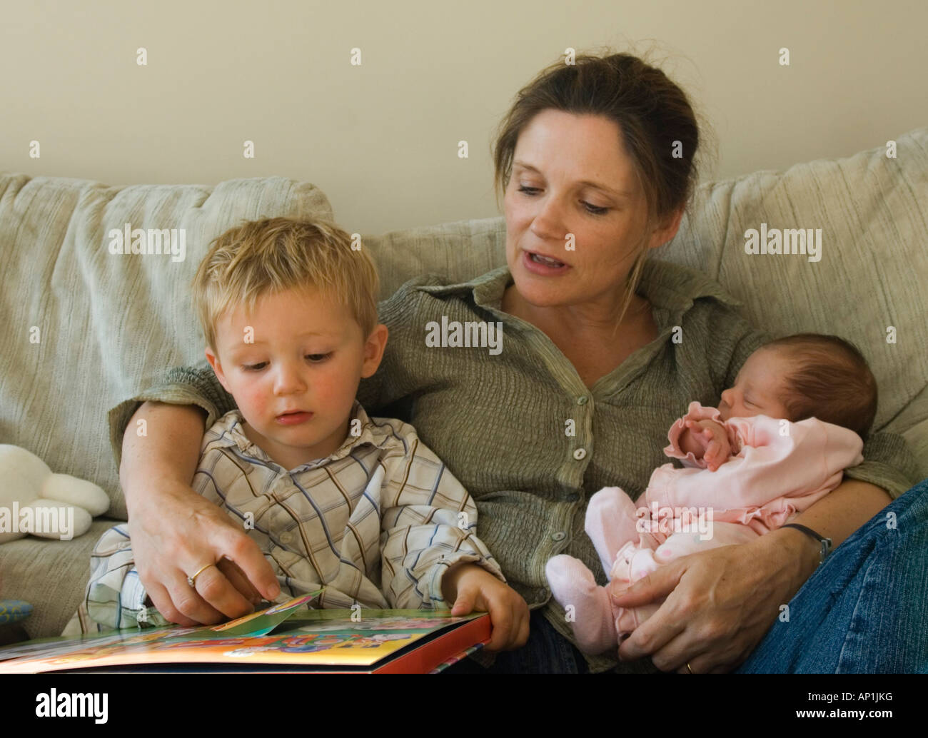 Mother toddler and new born girl reading childrens book on sofa at home ...