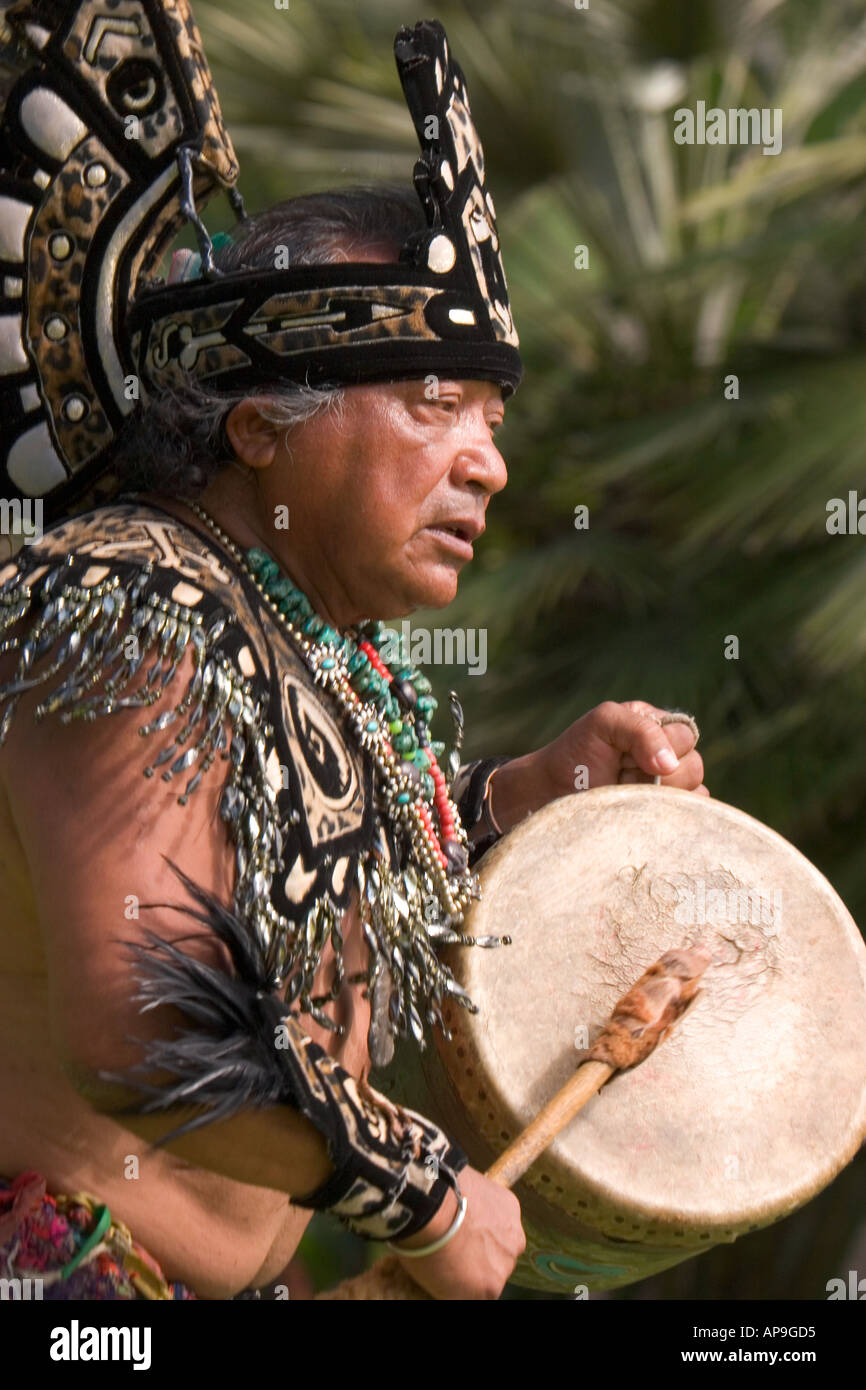 An Aztec man wearing traditional regalia, chanting and beating a drum ...