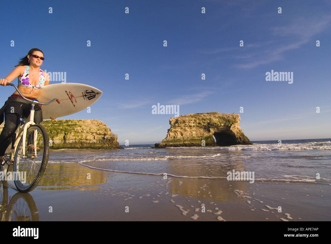 A woman with a surfboard riding a cruiser bike on the beach at Natural ...