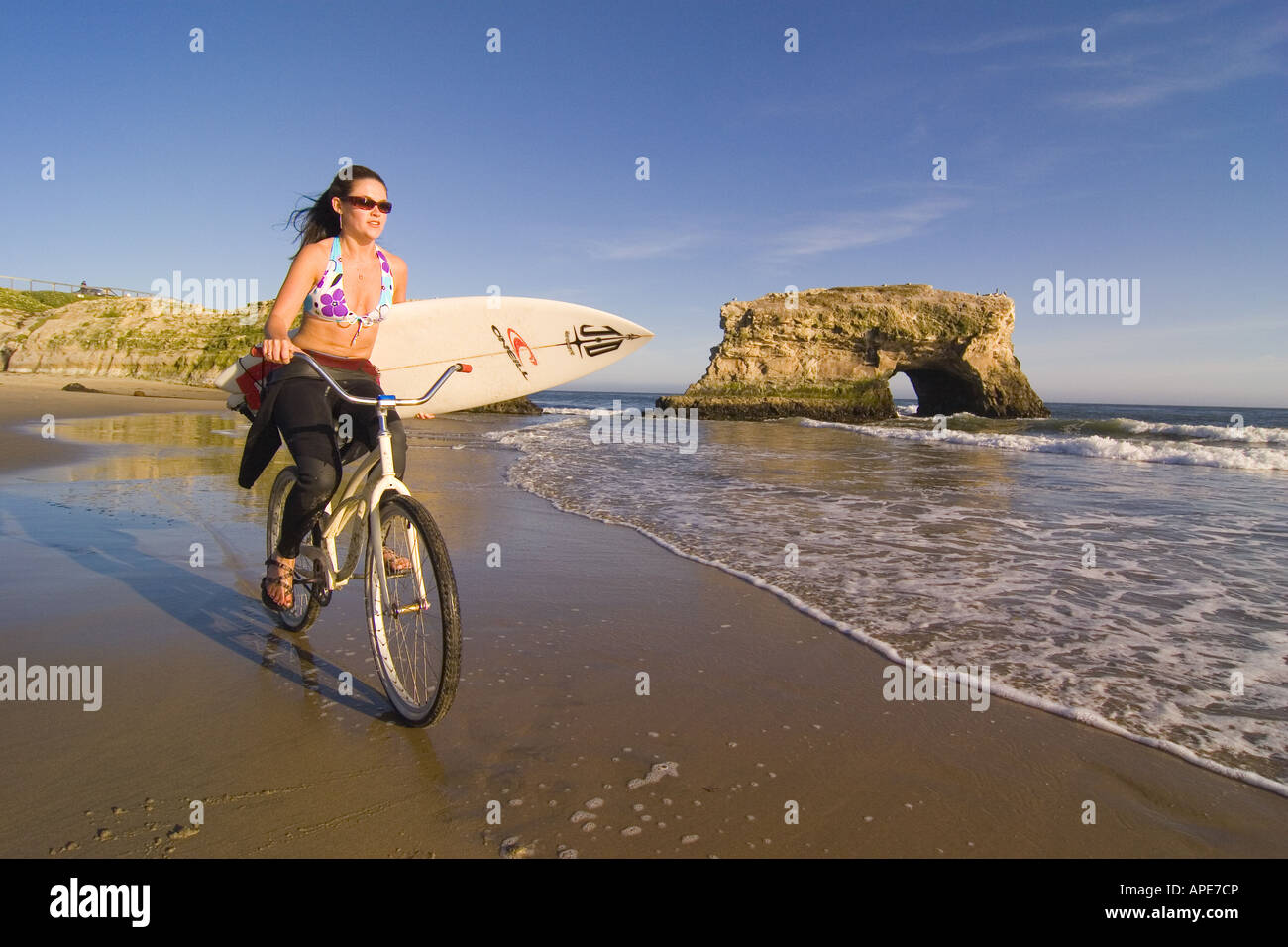 A woman with a surfboard riding a cruiser bike on the beach at Natural ...