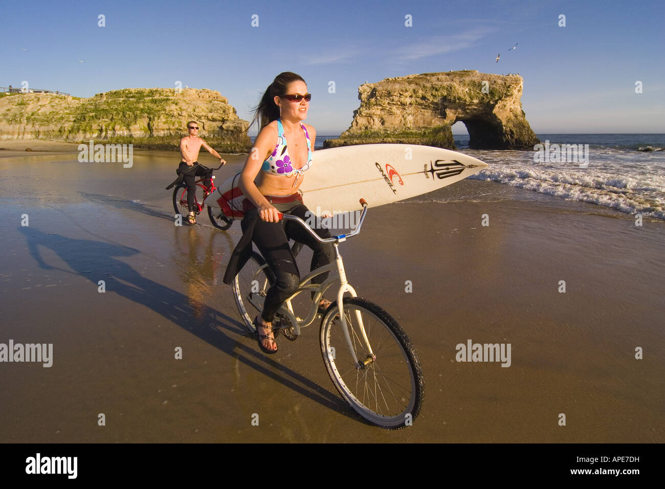 A woman and a man with a surfboard riding a cruiser bikes on the beach ...