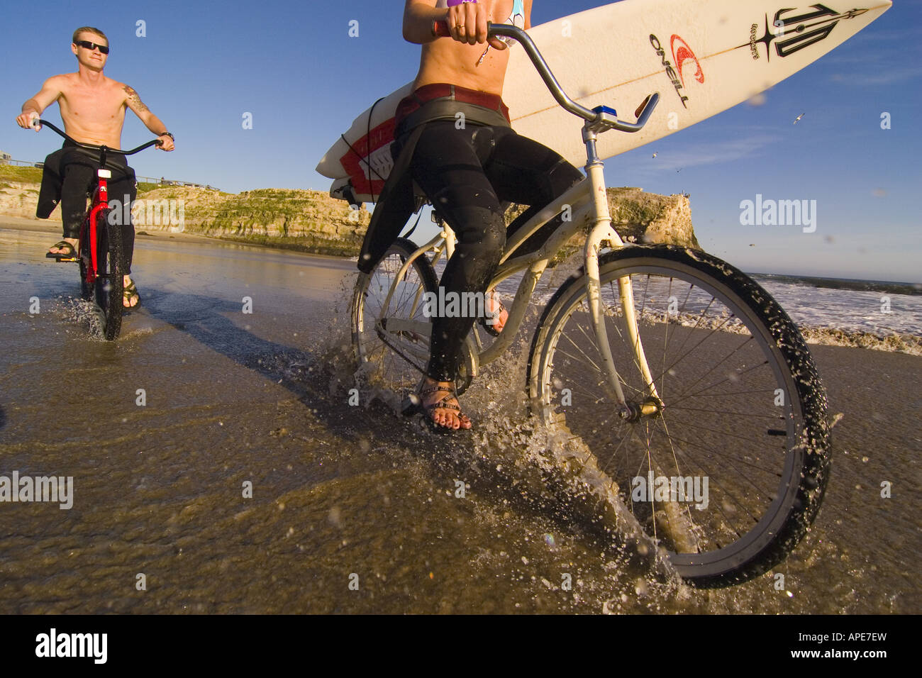 A woman and a man with a surfboard riding a cruiser bikes on the beach ...