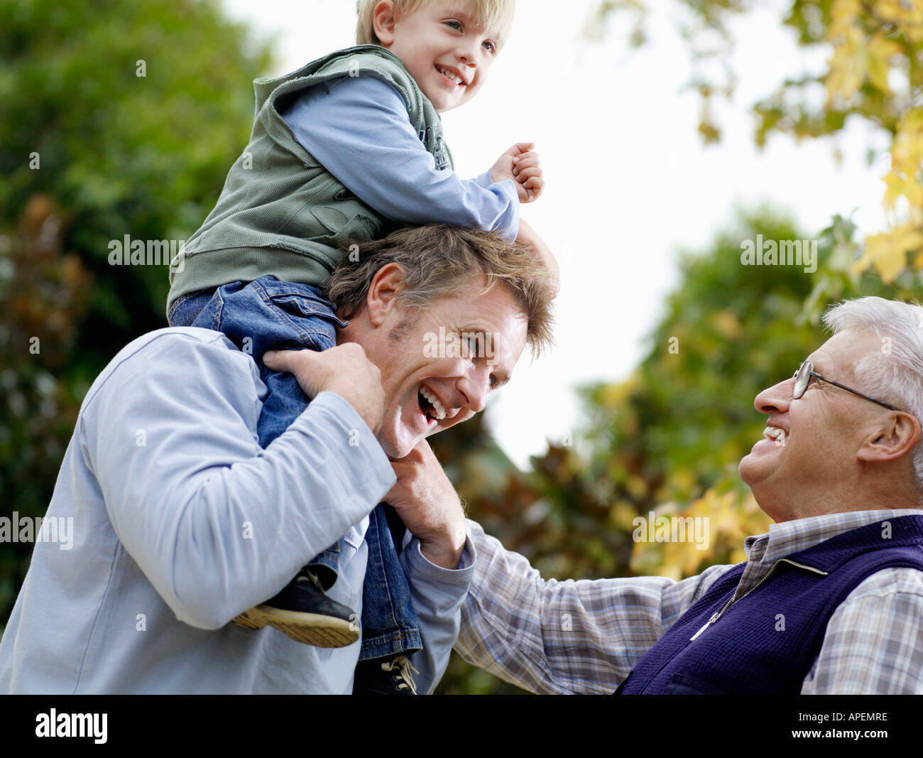 Boy (3-4) riding on father's shoulders, grandfather looking on, in yard ...