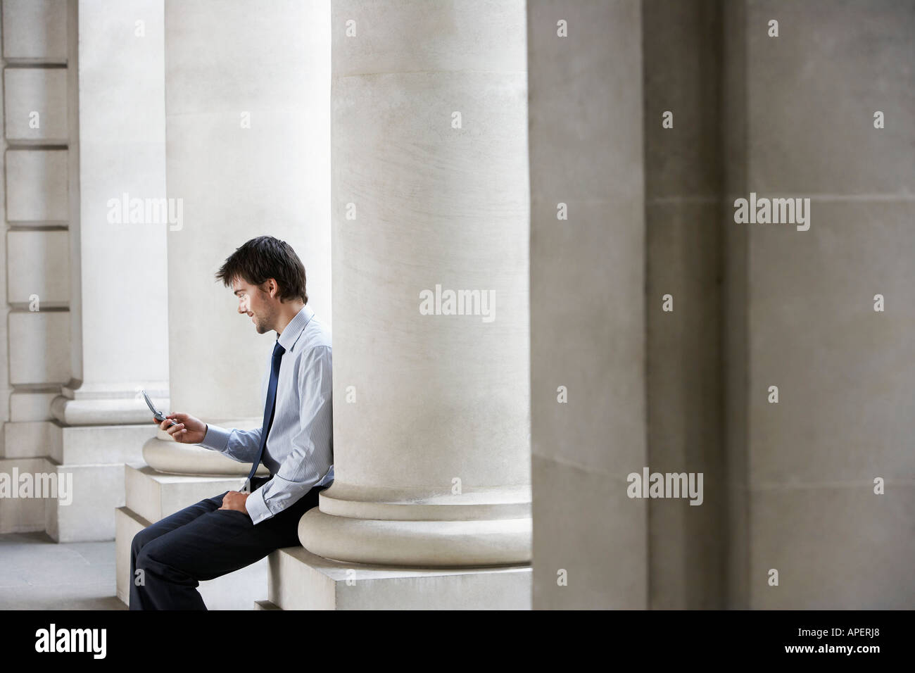 Businessman outside building sitting on pillar sending text message ...