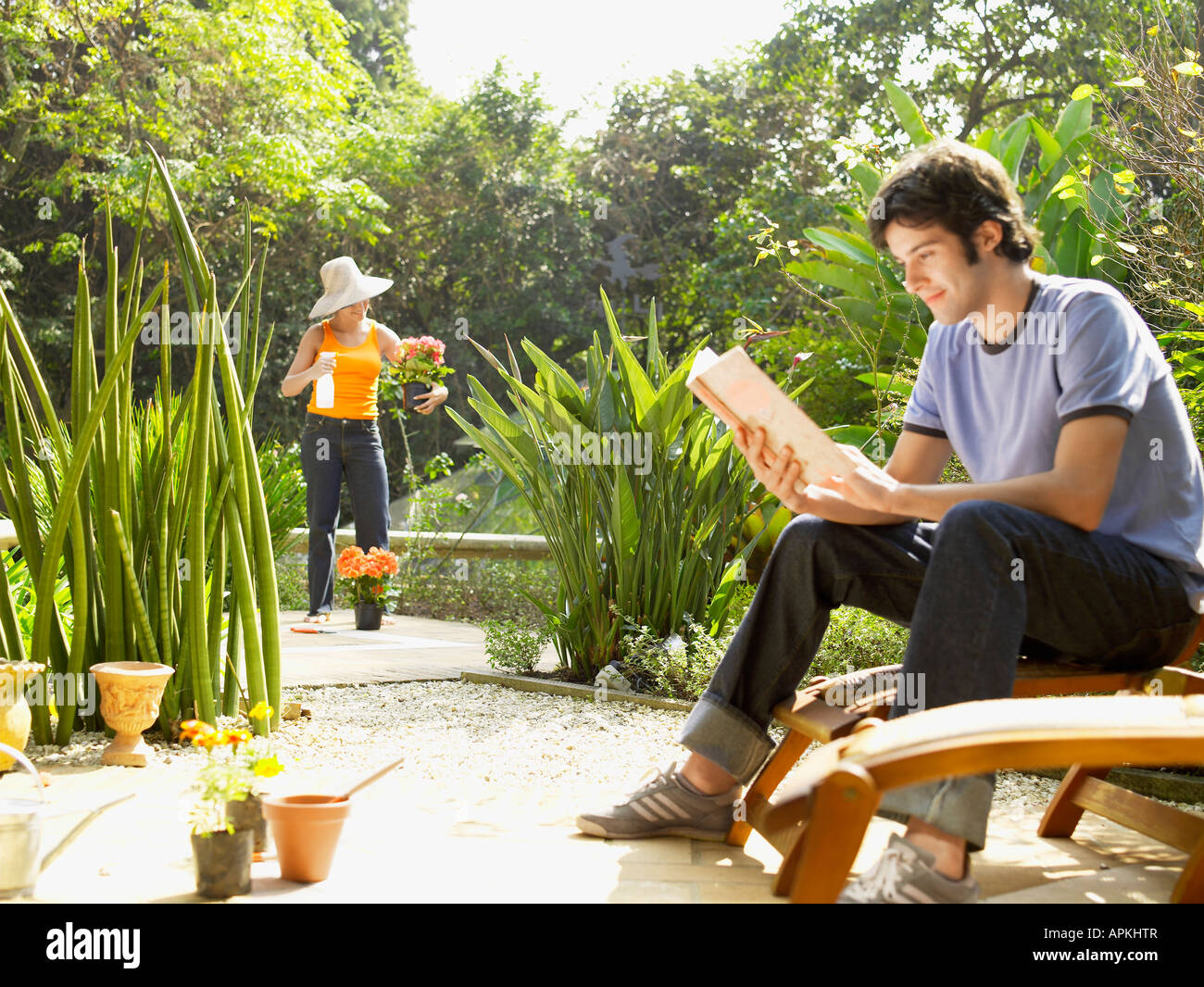 Man reading book with woman gardening in background Stock Photo - Alamy