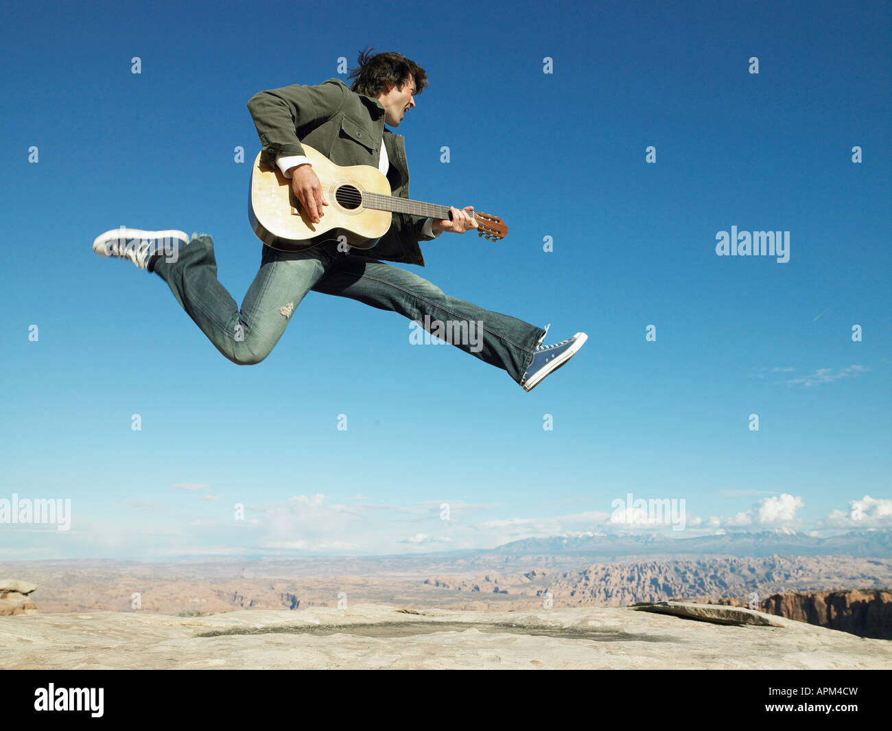 Mid adult man jumping and playing acoustic guitar, rock strata in ...