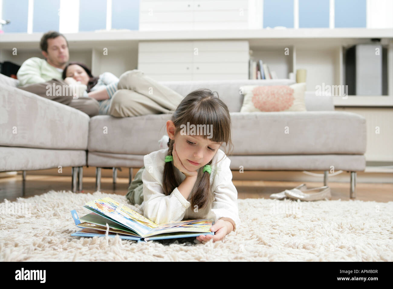 Girl reading book on floor, parents on sofa in background Stock Photo ...