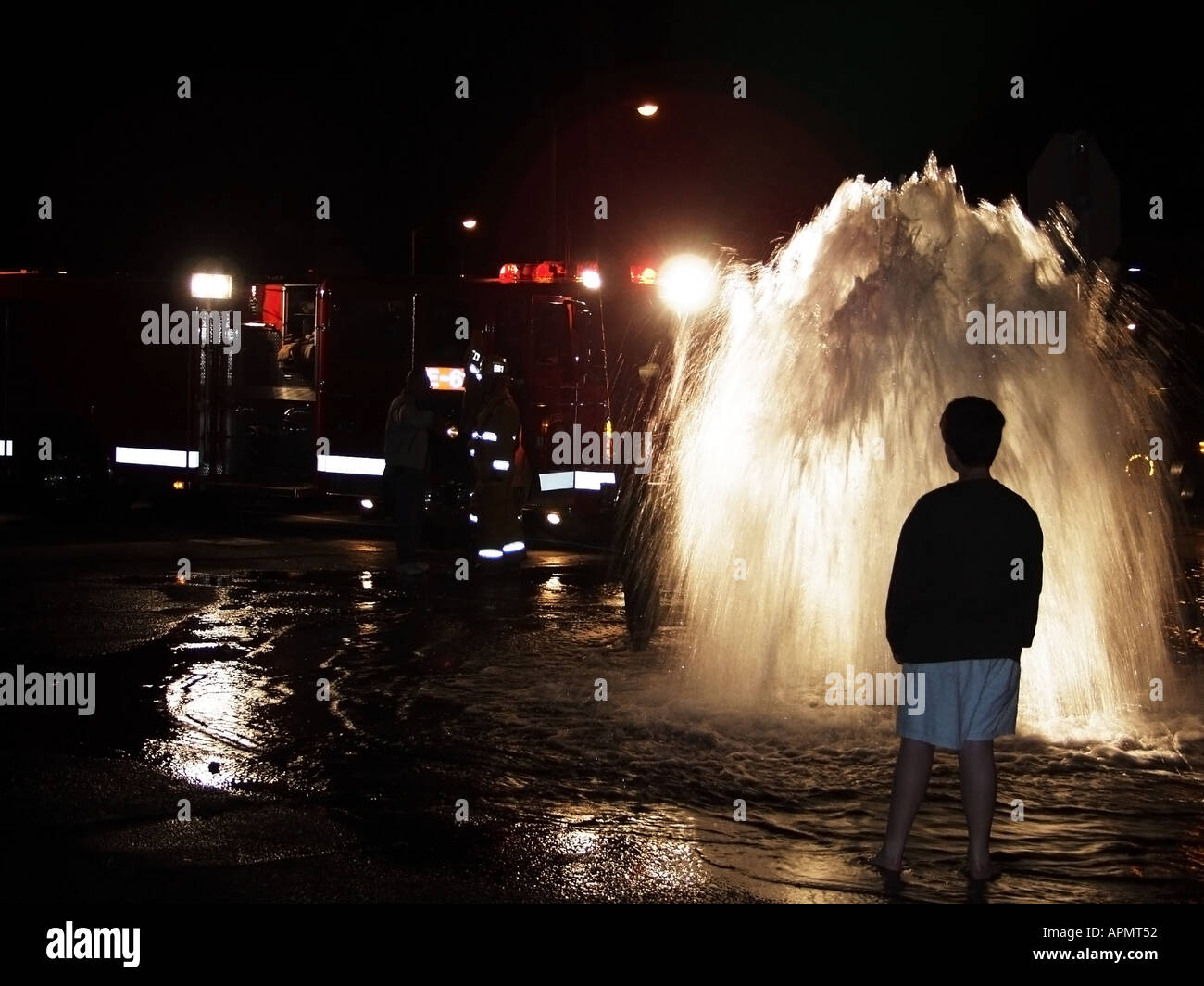 Young boy silhouetted against fire hydrant water geyser at night ...