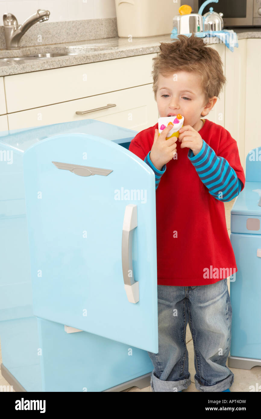 a boy plays in a kitchen with a toy fridge, he stands in its doorway ...