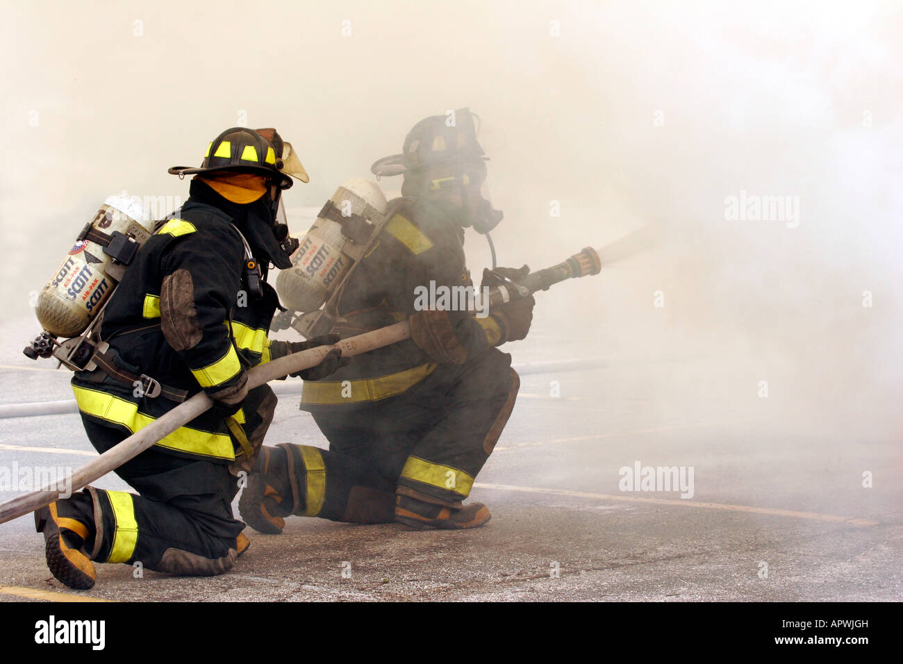 Two firefighters on a hoseline spraying water into a fire surrounded by ...