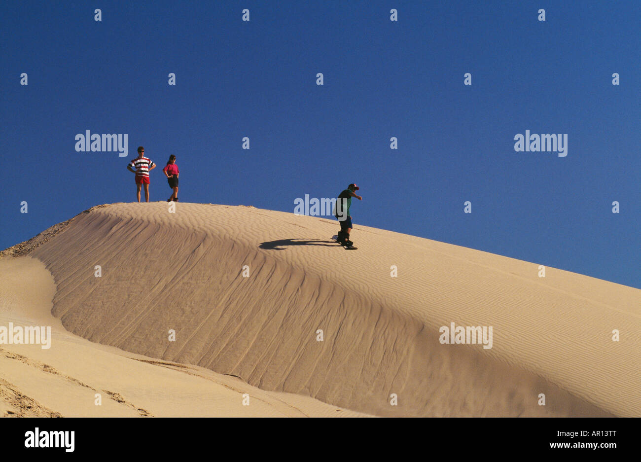 A man slides on the sand dune with a snowboard as a couple look at him ...