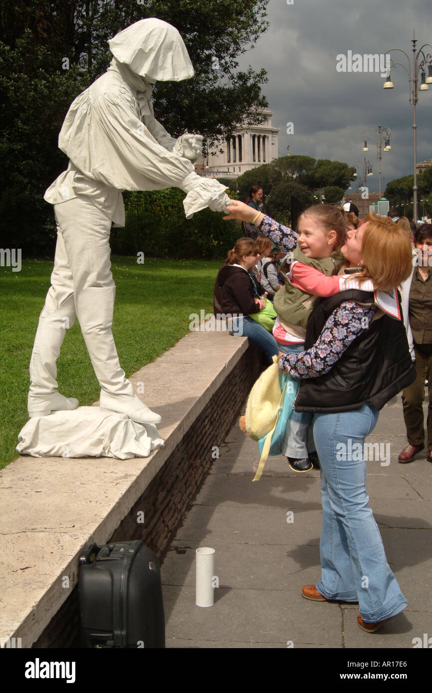 Mother and child reach out to white painted mime posing as a statue act ...